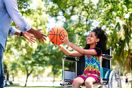 Djevojčica u invalidskim kolicima koja se zabavlja sa ocem dok zajedno igra košarku u parku