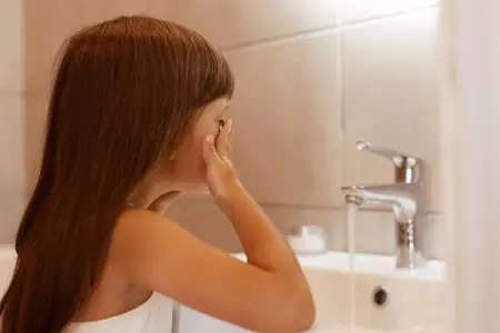 Tamsiaplaukės moters, stovinčios vonios kambaryje ir praususios veidą po pabudimo arba prieš einant miegoti, nuotrauka. Higienos procedūros namuose