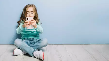 Портрет маленькой девочки, сидящей на деревянном полу и смотрящей на смартфон